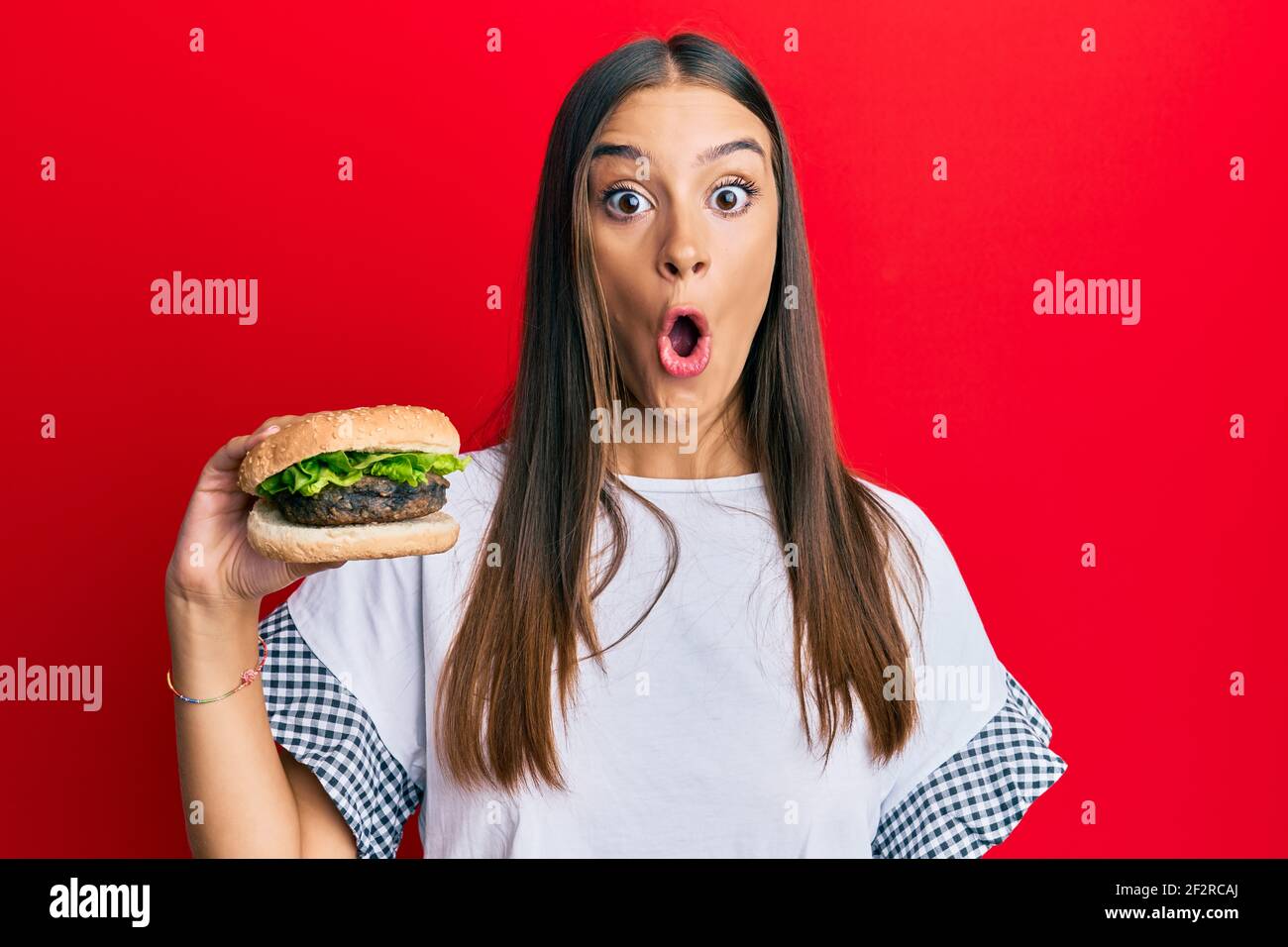 Young hispanic woman eating hamburger scared and amazed with open mouth ...