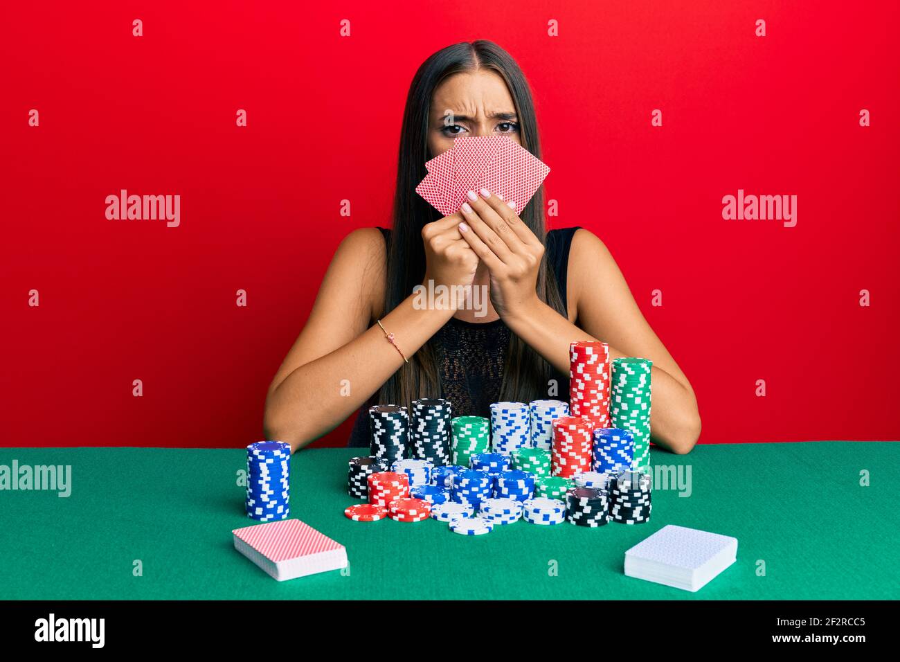 Young hispanic woman playing gambling poker covering face with cards ...