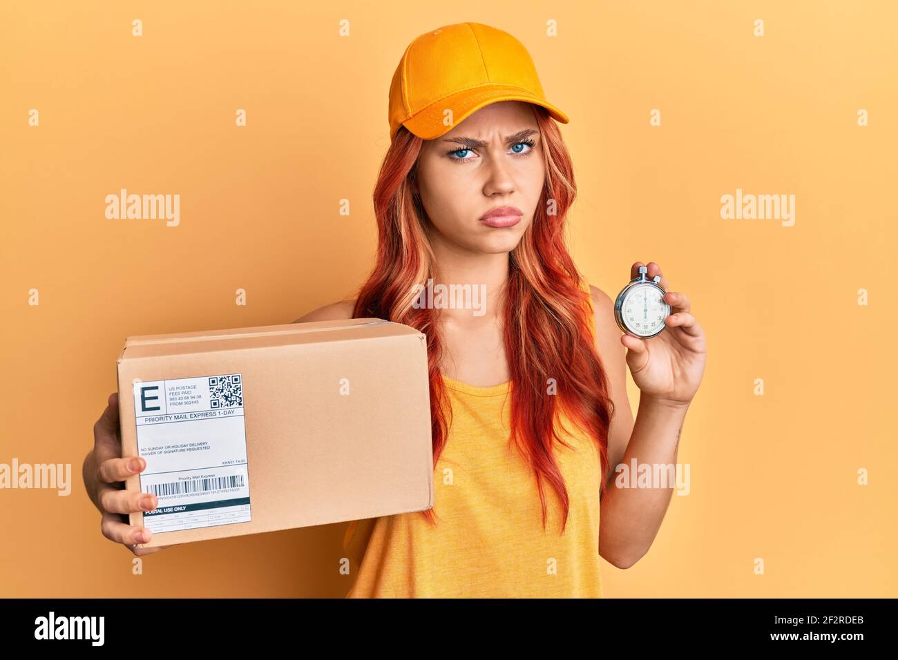 Young redhead woman holding delivery package and stopwatch skeptic and ...