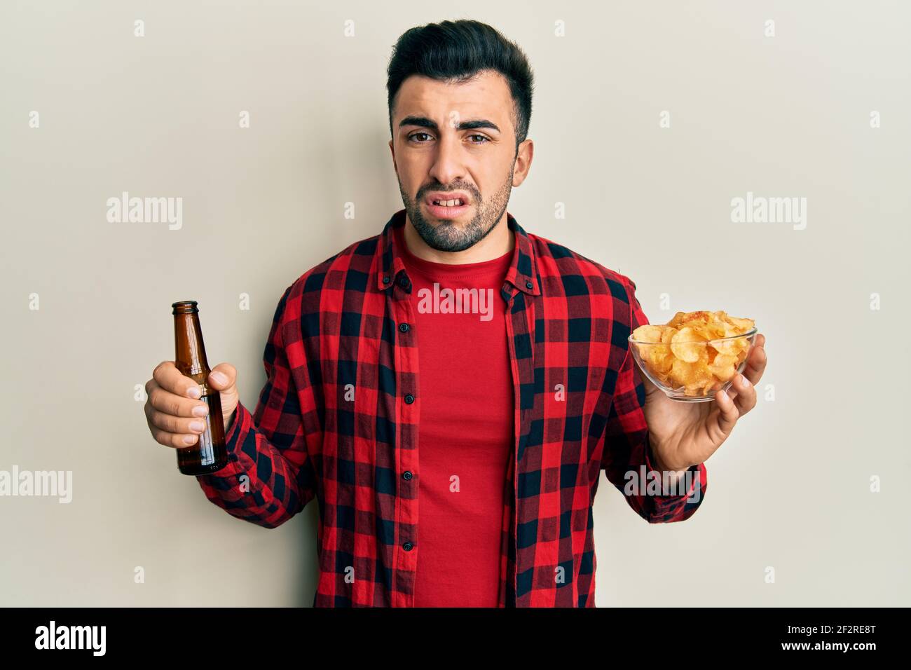 Young hispanic man drinking a bottle of beer and eating chip potatoes ...