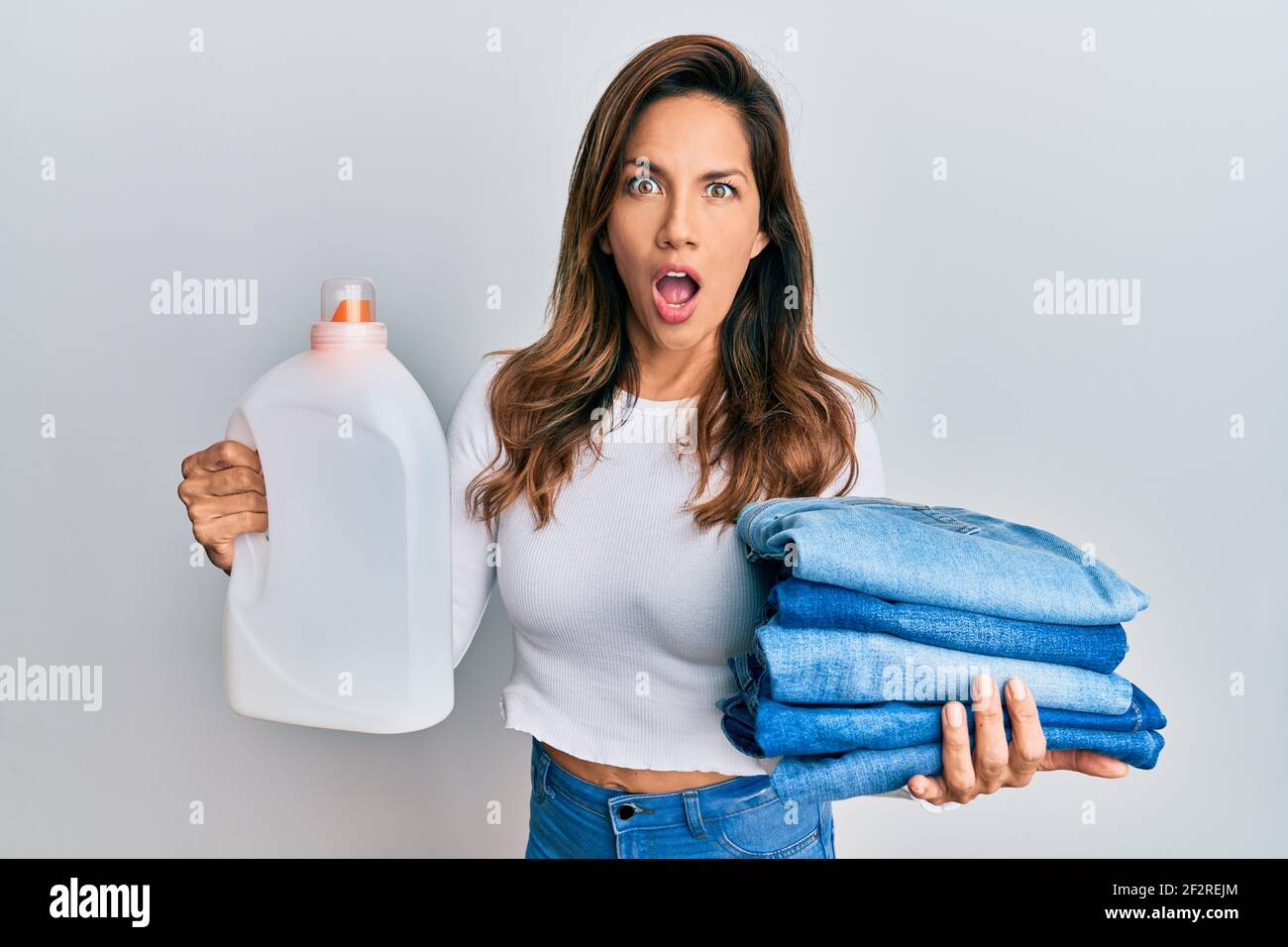 Young latin woman holding jeans for laundry and detergent bottle afraid and shocked with ...