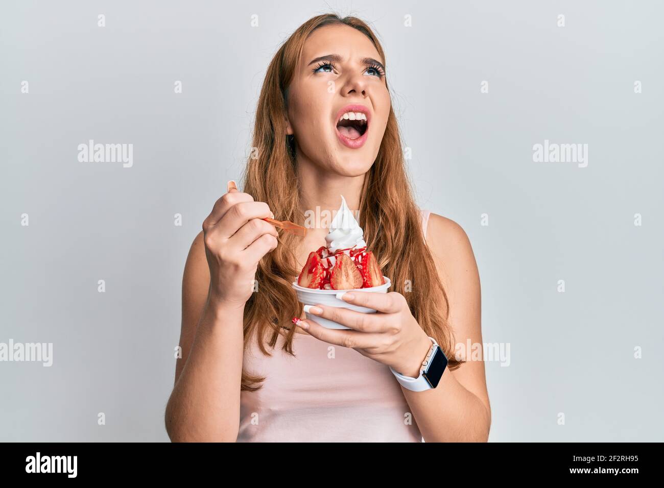 Young blonde woman eating strawberry ice cream angry and mad screaming ...