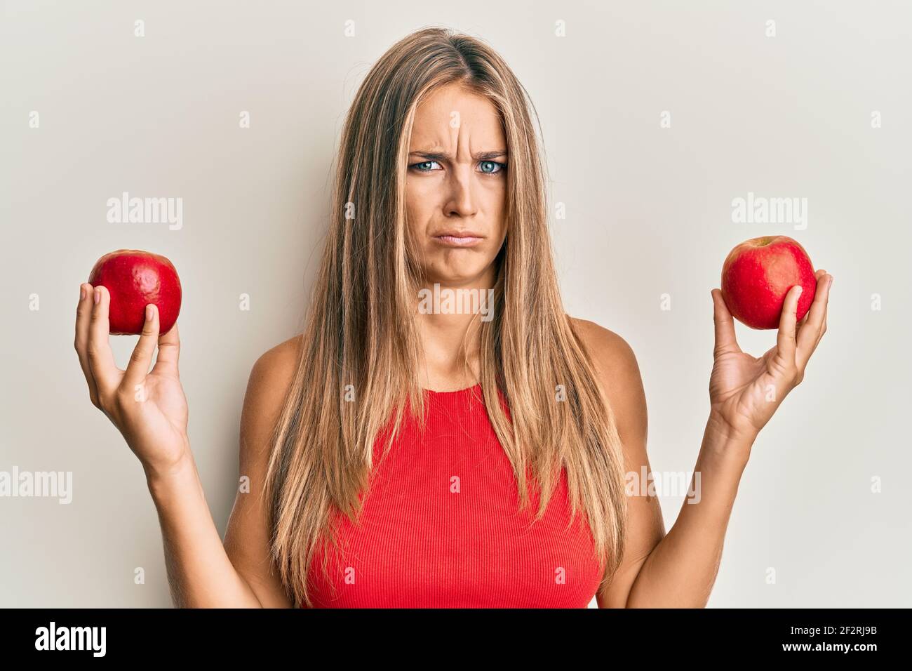 Young blonde woman holding red apples skeptic and nervous, frowning ...