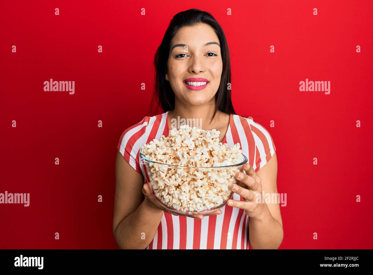 Young latin woman holding popcorn looking positive and happy standing ...