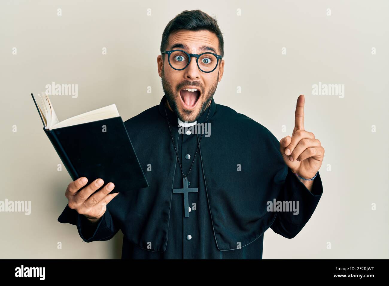 Young hispanic priest man holding bible with finger up celebrating ...