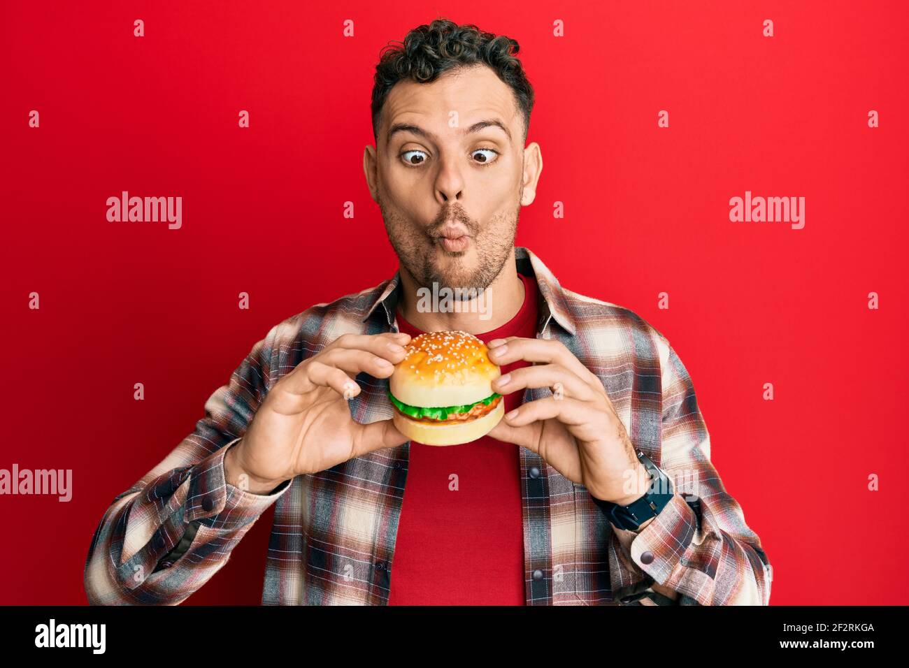 Young hispanic man eating a tasty classic burger making fish face with ...