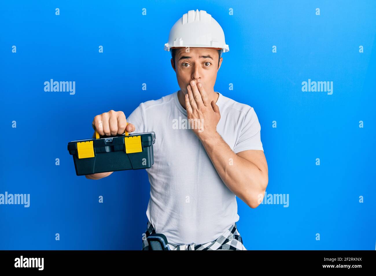Handsome young man wearing safety helmet holding toolbox covering mouth ...