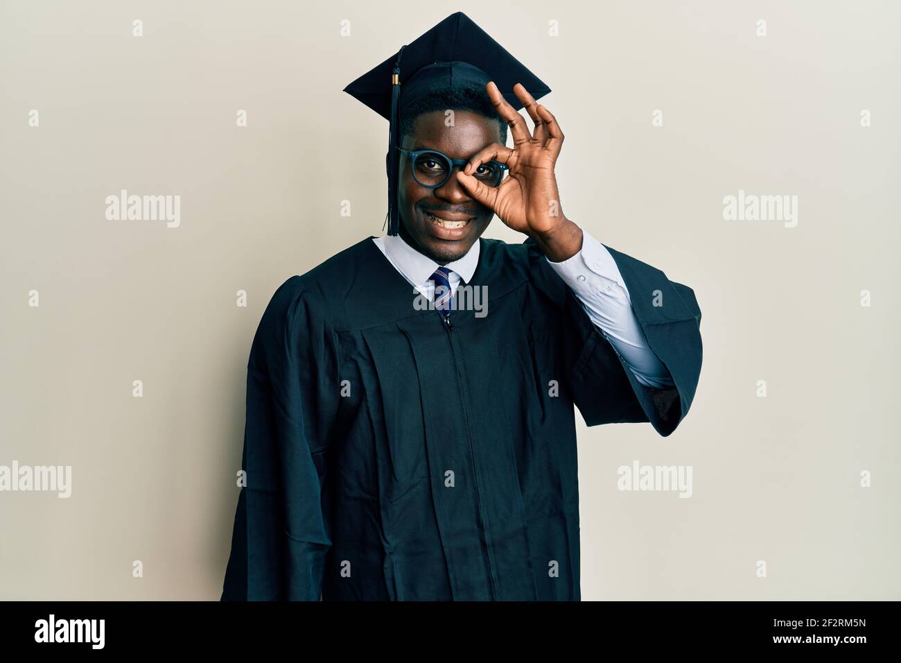 Handsome black man wearing graduation cap and ceremony robe doing ok gesture with hand smiling ...