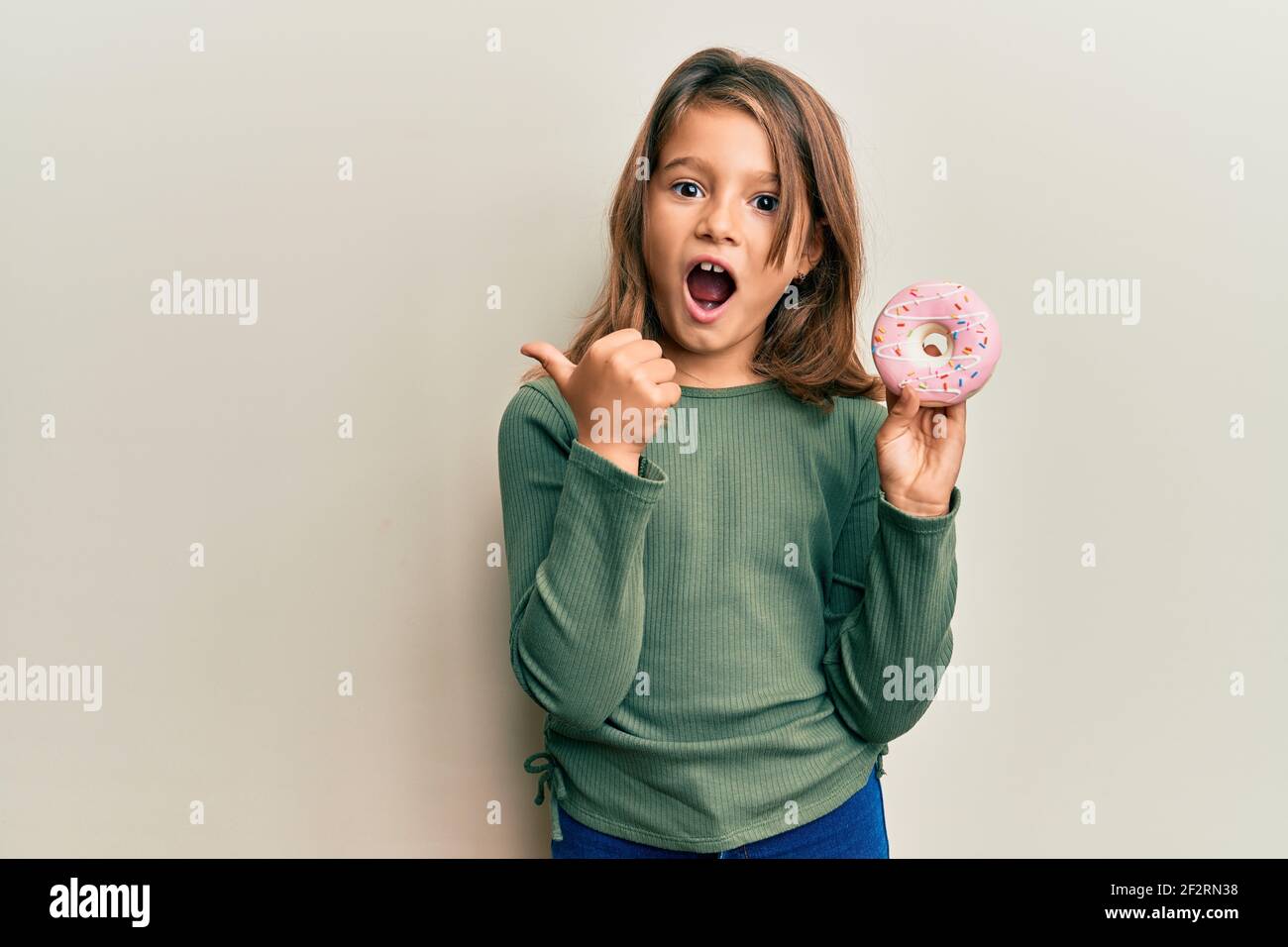 Little beautiful girl holding tasty colorful doughnut pointing thumb up ...