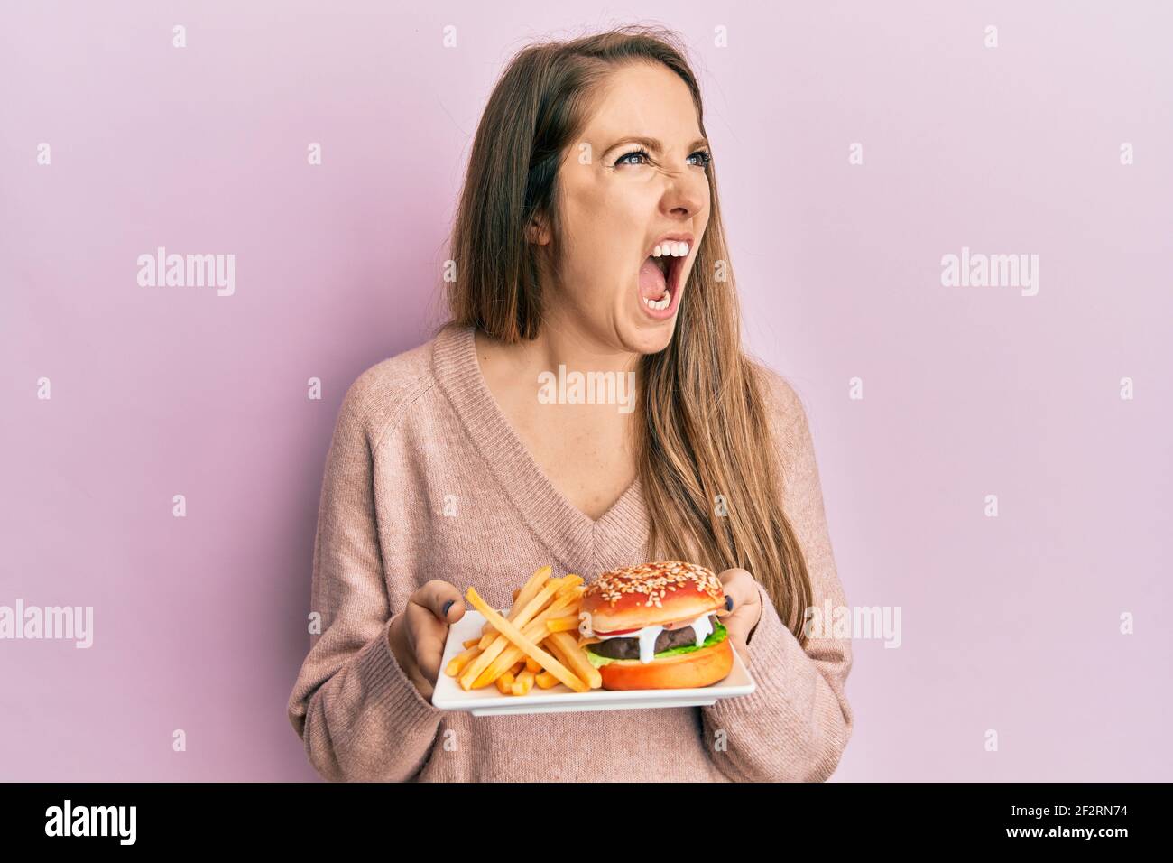 Young blonde woman eating a tasty classic burger with fries angry and ...