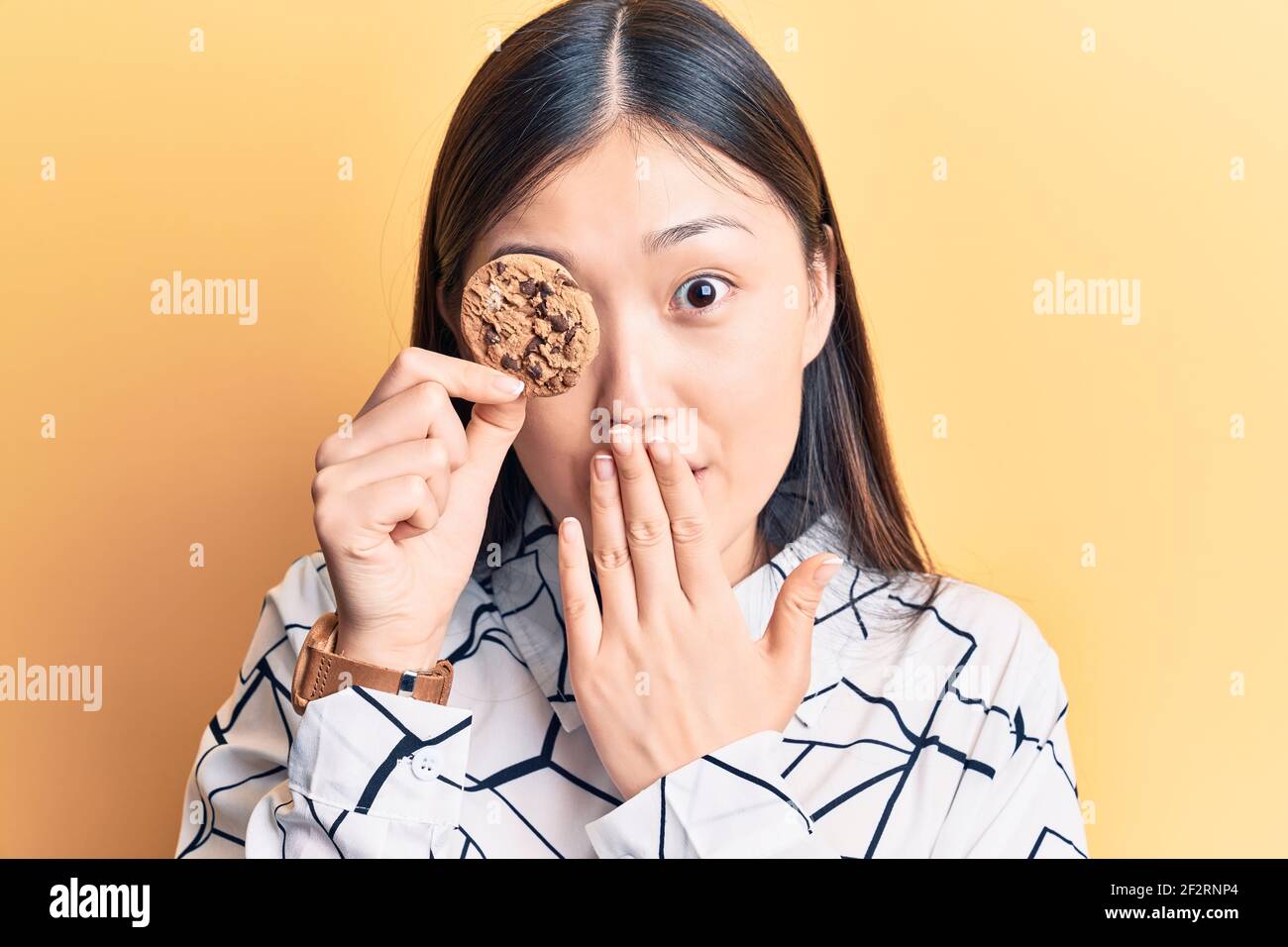 Young beautiful chinese woman holding cookie over eye covering mouth ...