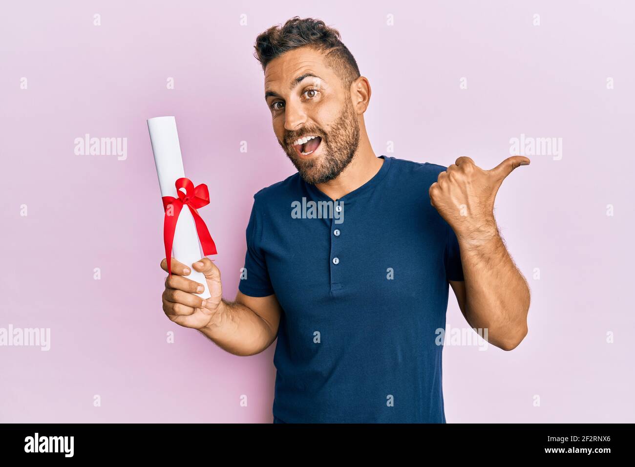 Handsome man with beard holding graduate degree diploma pointing thumb ...