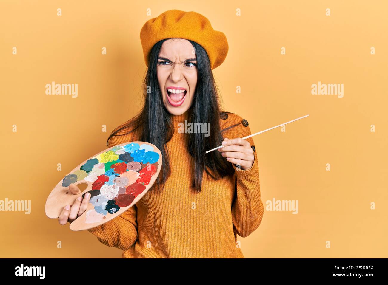 Young brunette woman holding paintbrush and palette wearing beret angry ...