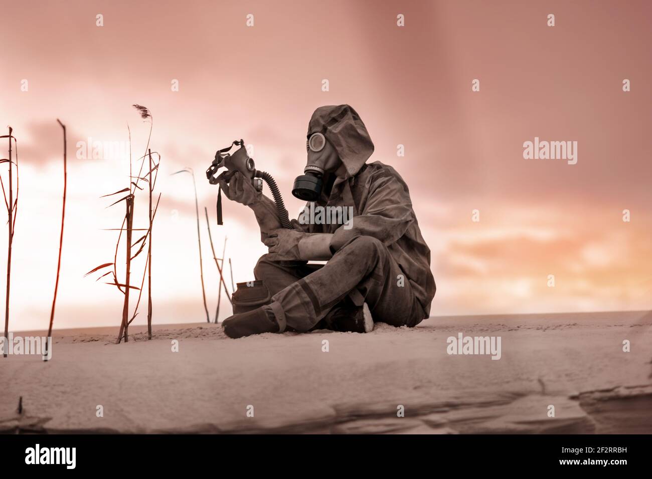 A man in a gas mask and protective suit sits on the sand and holds the ...