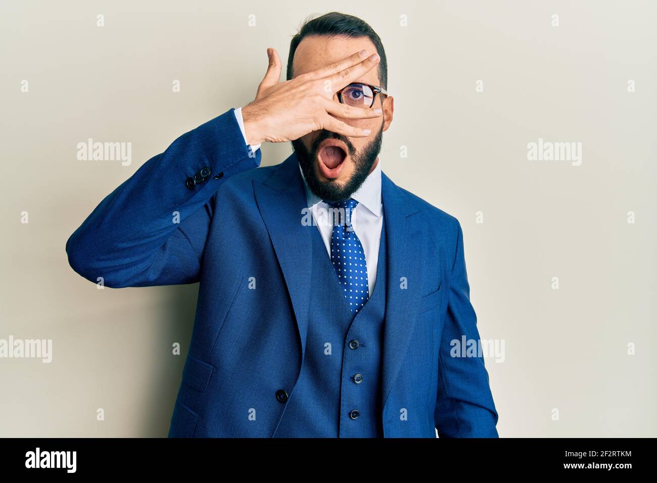 Young man with beard wearing business suit and tie peeking in shock ...