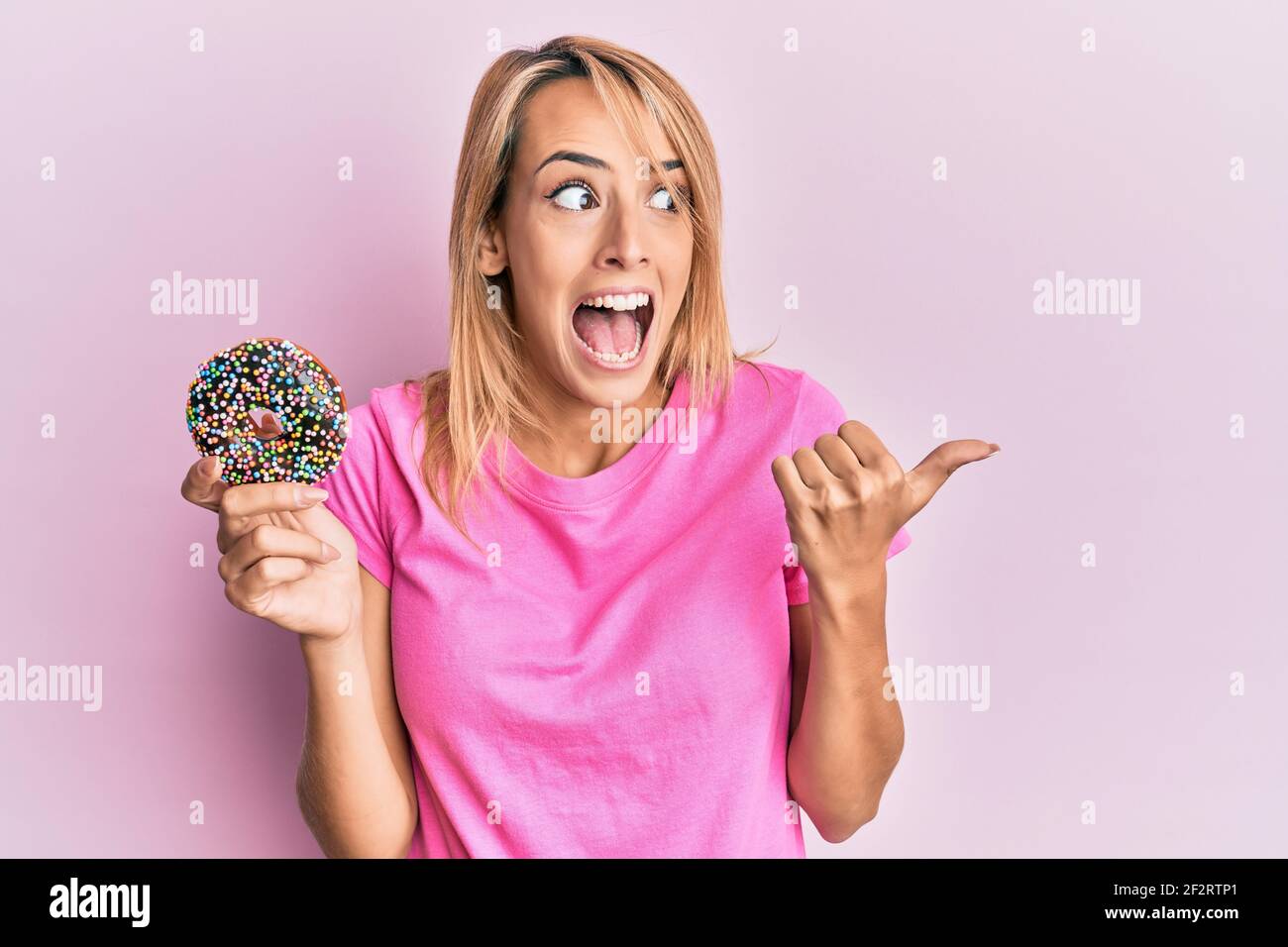 Beautiful blonde woman holding donut pointing thumb up to the side ...