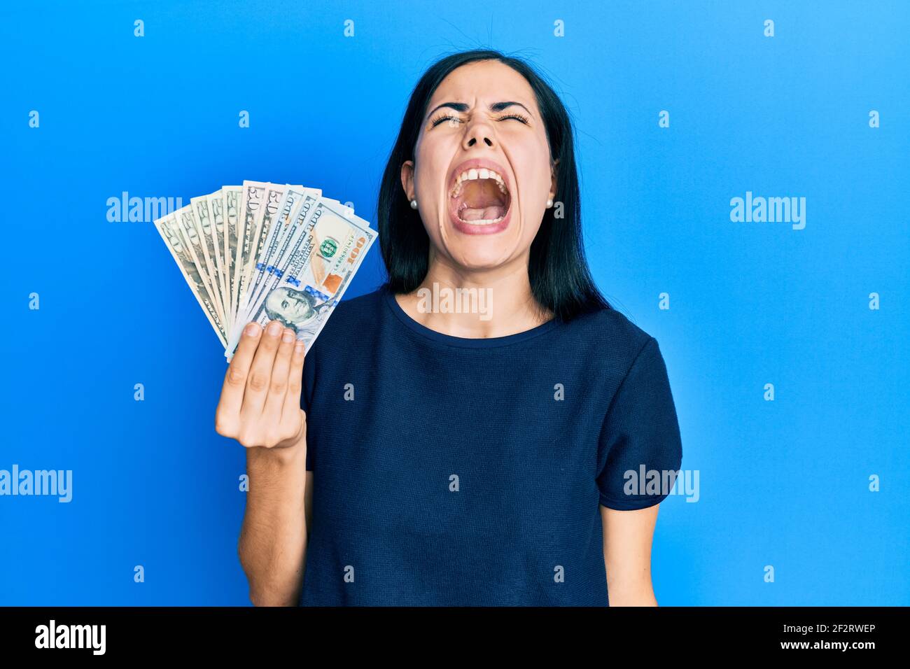 Beautiful young woman holding dollars angry and mad screaming ...