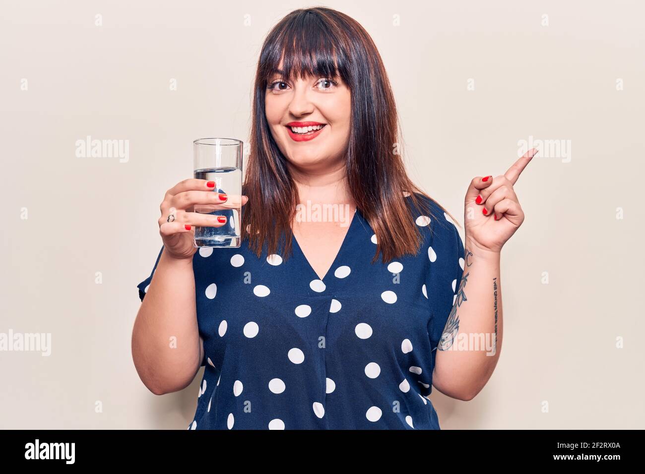 Young plus size woman drinking glass of water smiling happy pointing ...