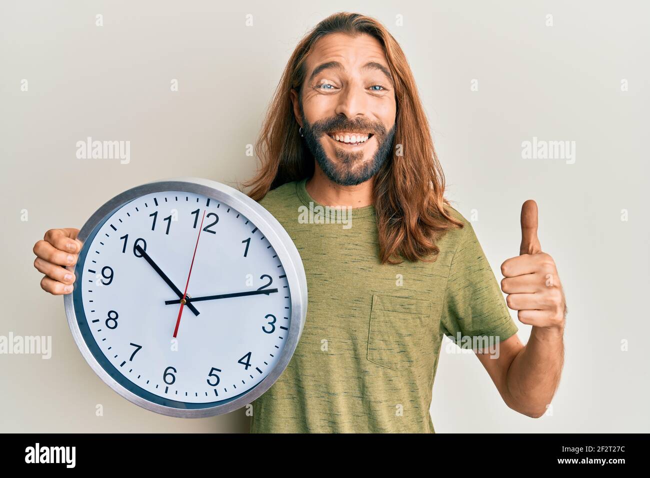 Attractive man with long hair and beard holding big clock smiling happy ...