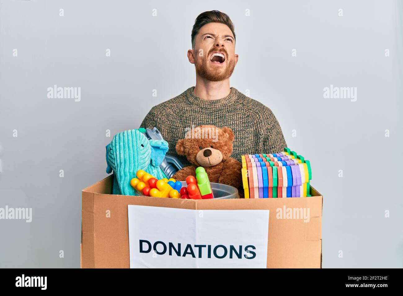 Young redhead man holding donation box with toys angry and mad ...
