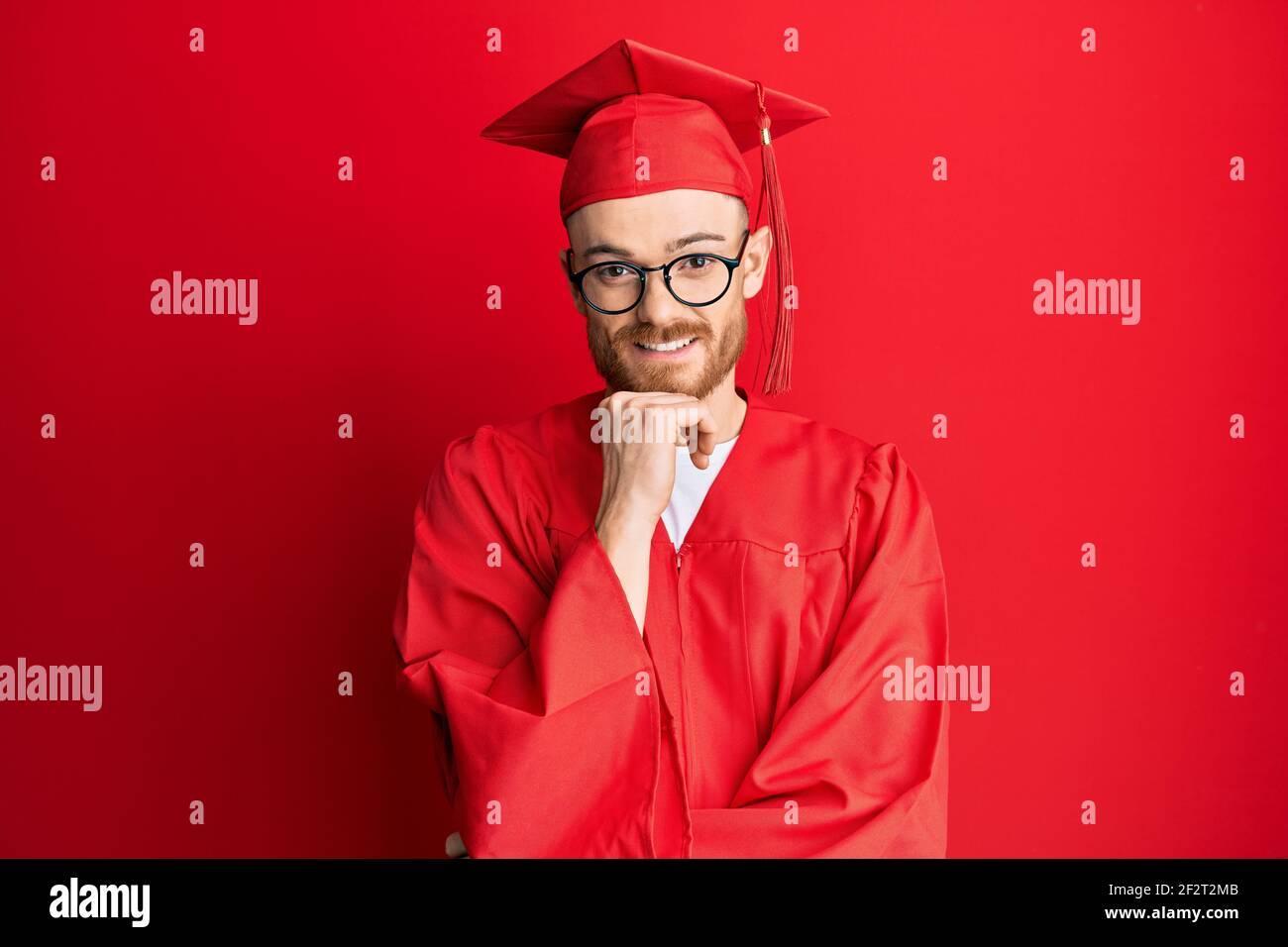 Young redhead man wearing red graduation cap and ceremony robe looking ...