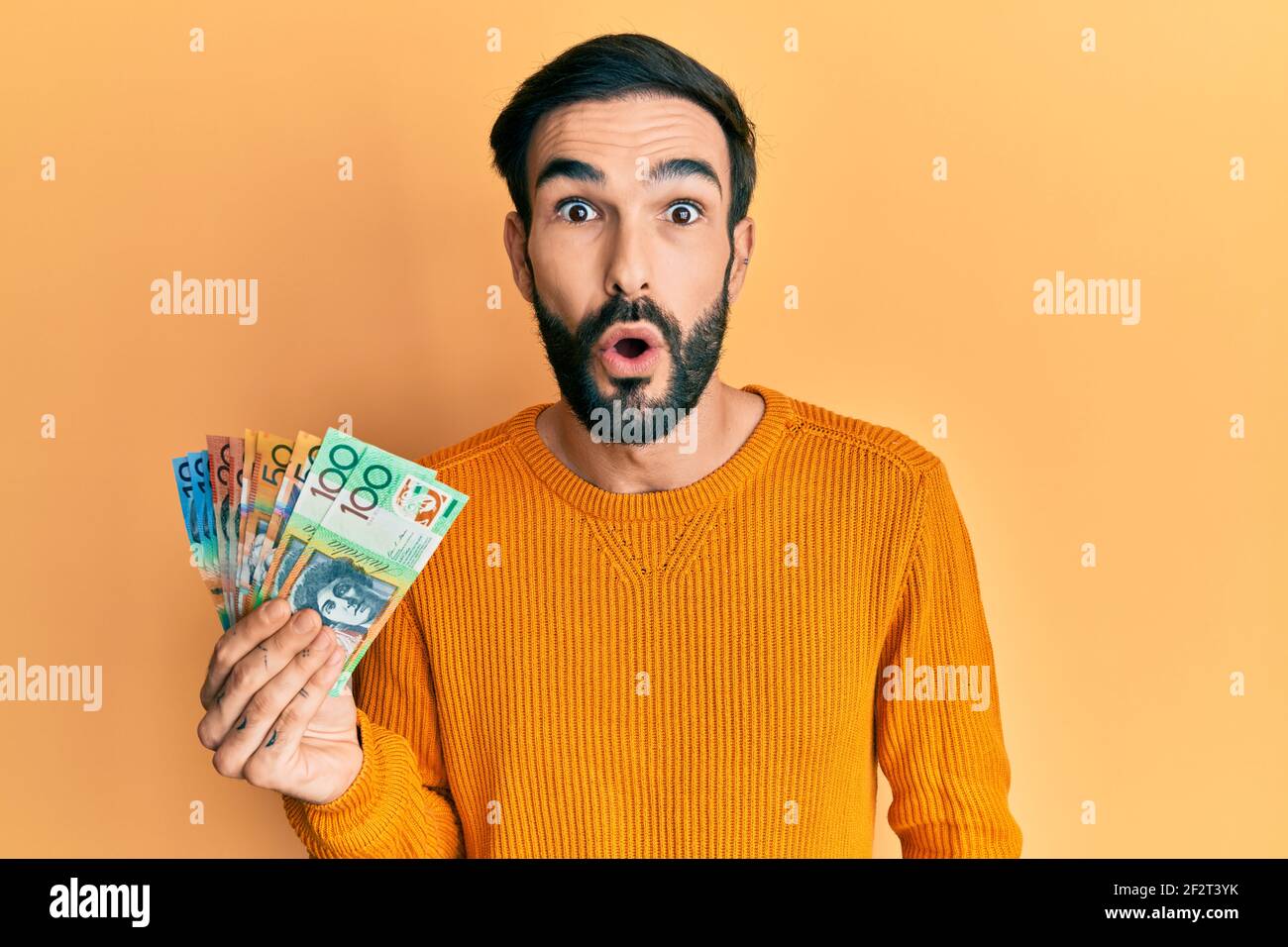 Young hispanic man holding australian dollars scared and amazed with ...