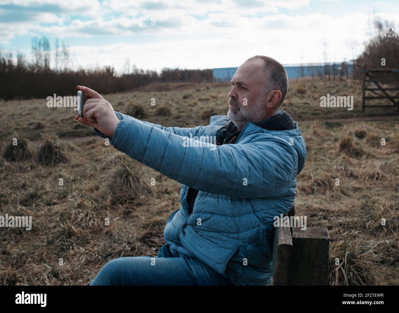 old man in a blue coat with the mobile phone sitting on the bench in a ...