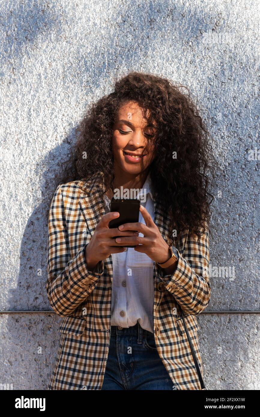 African American female in trendy outfit with curly hair browsing phone ...