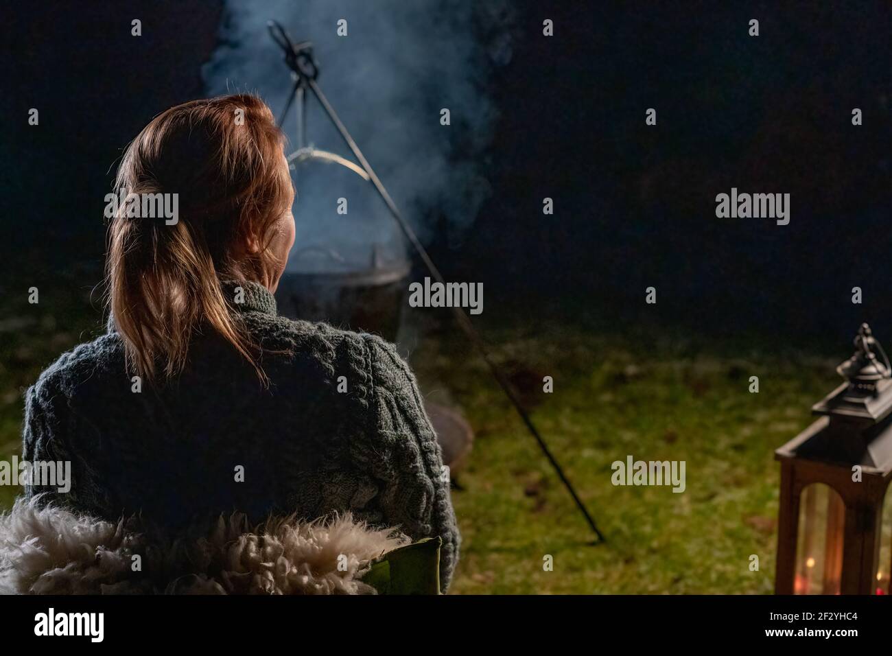 Young blond woman sits outside by the campfire in the woods. It is dark ...