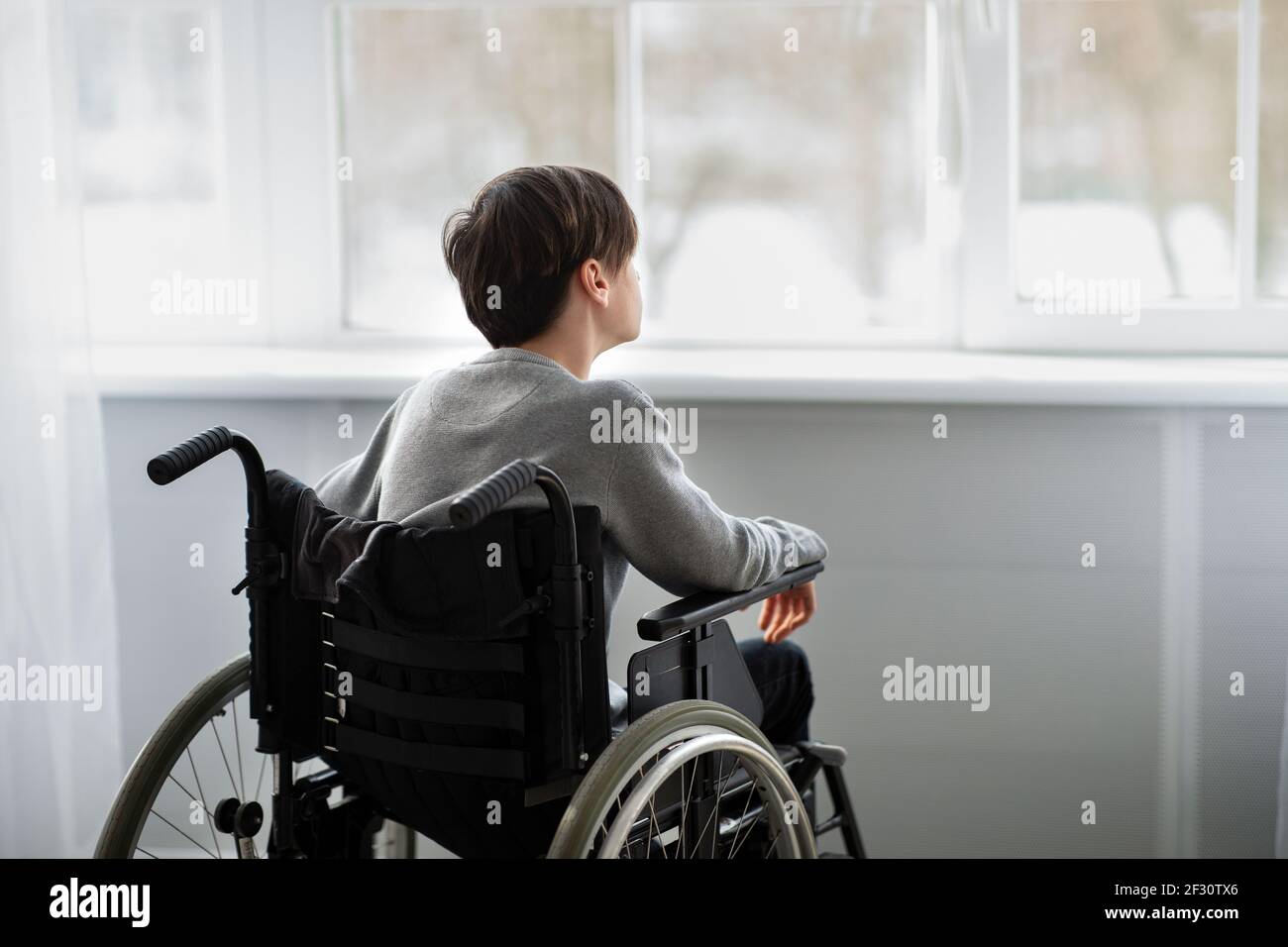 Back view of disabled teenager in wheelchair looking out window, having ...
