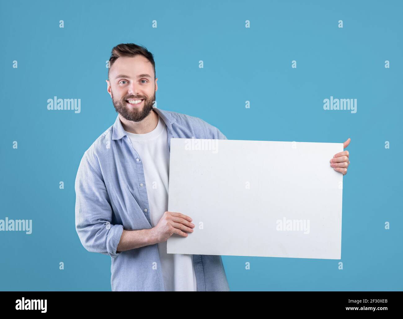 Joyful Caucasian man demonstrating white blank placard on blue studio ...