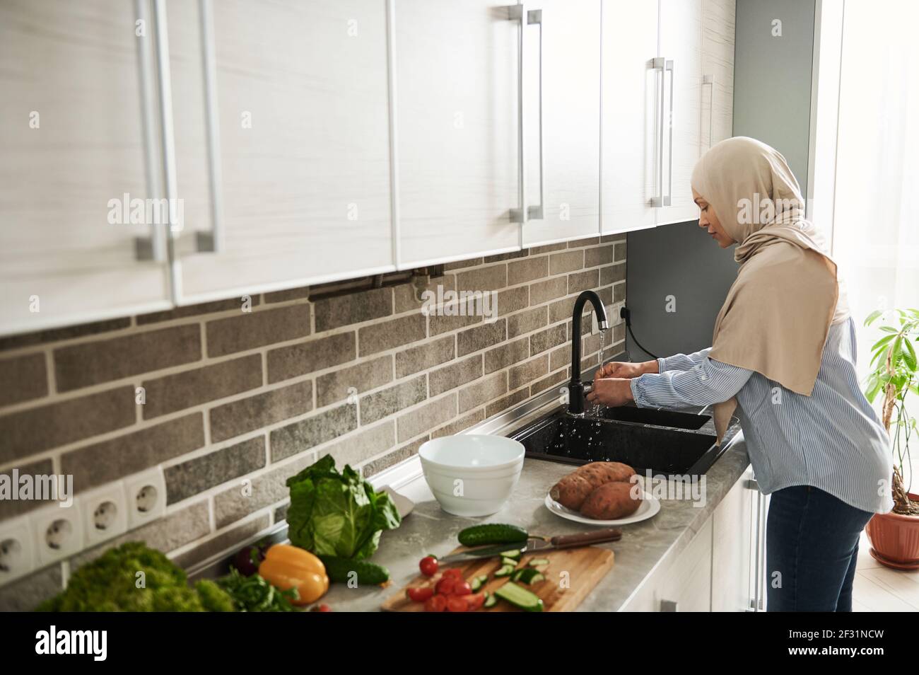 Muslim woman with covered head washing vegetable leaf in the kitchen ...
