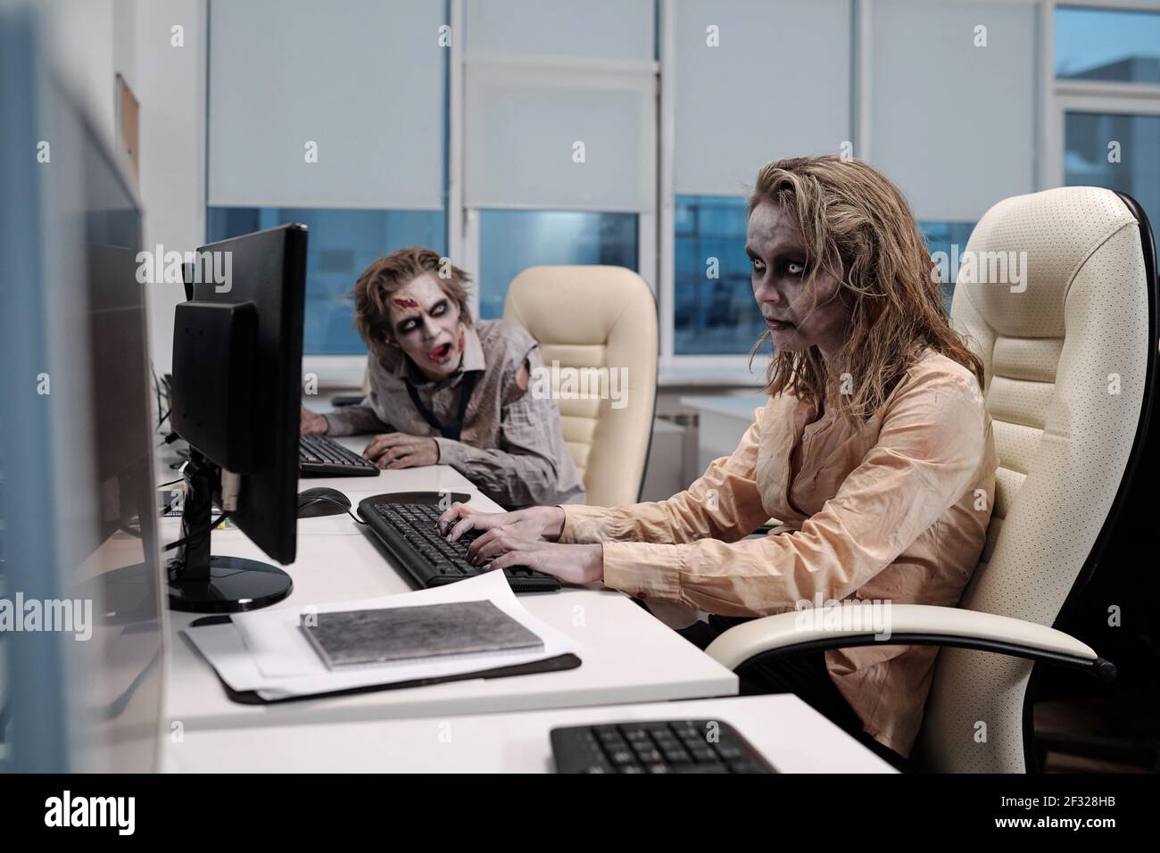 Young spooky zombie businesswoman sitting by desk in front of computer ...