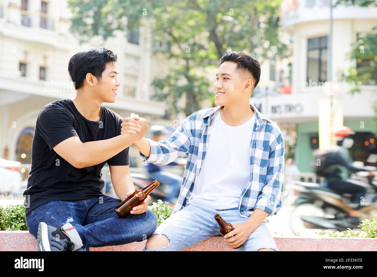 Asian young man sitting on bench with his friend and drinking beer ...