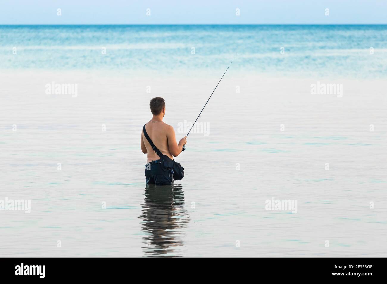 A young guy without a shirt stands knee-deep in the water of the sea in ...