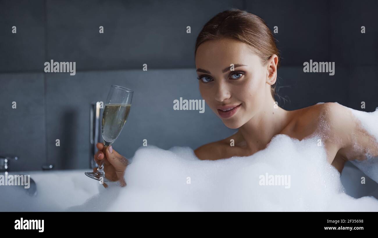 Positive Woman Taking Bath With Foam And Holding Glass Of Champagne In