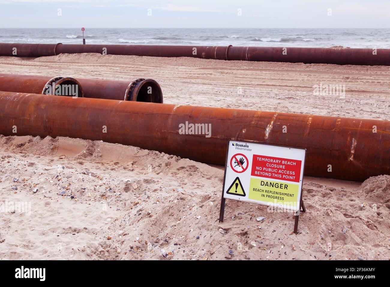 Sign warning of danger and beach closure. Large metal pipes or pipeline ...