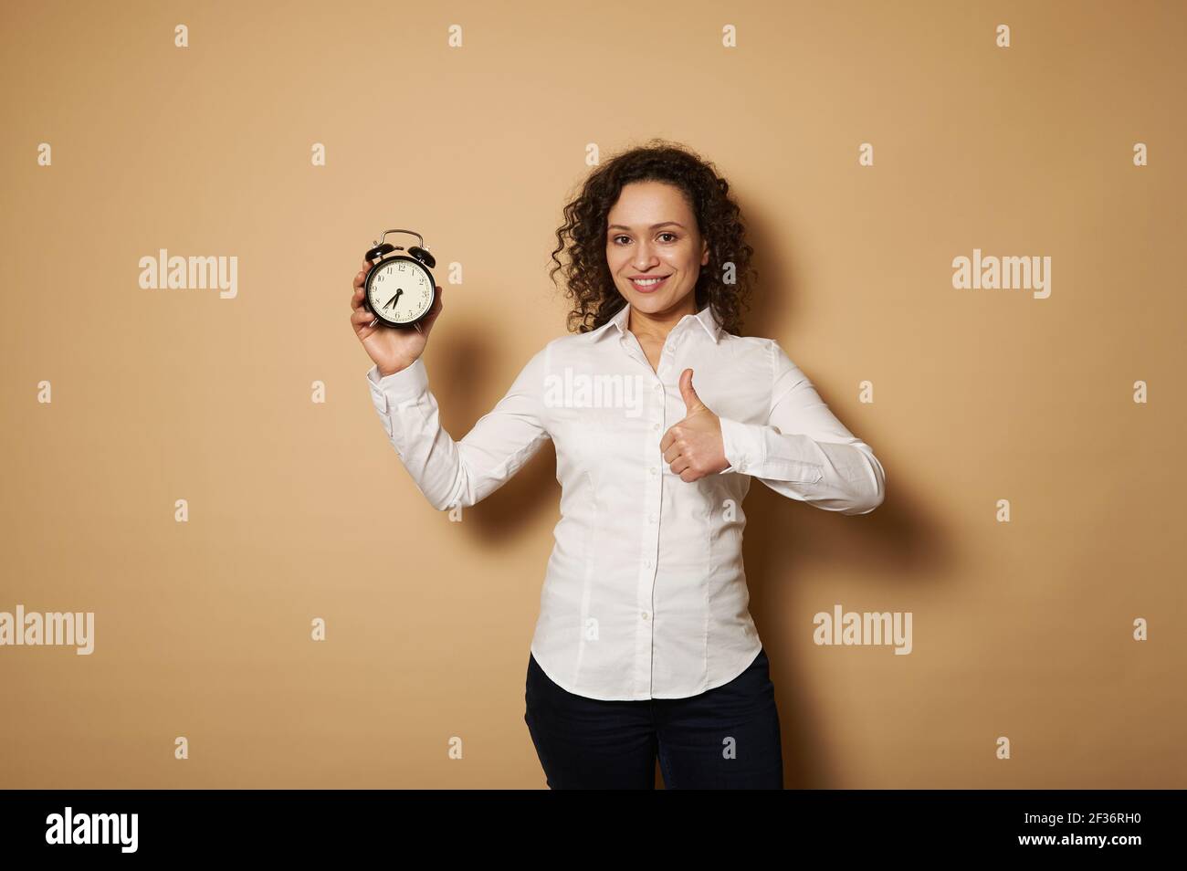 Young curly woman holds an alarm clock and shows thumb up while posing ...