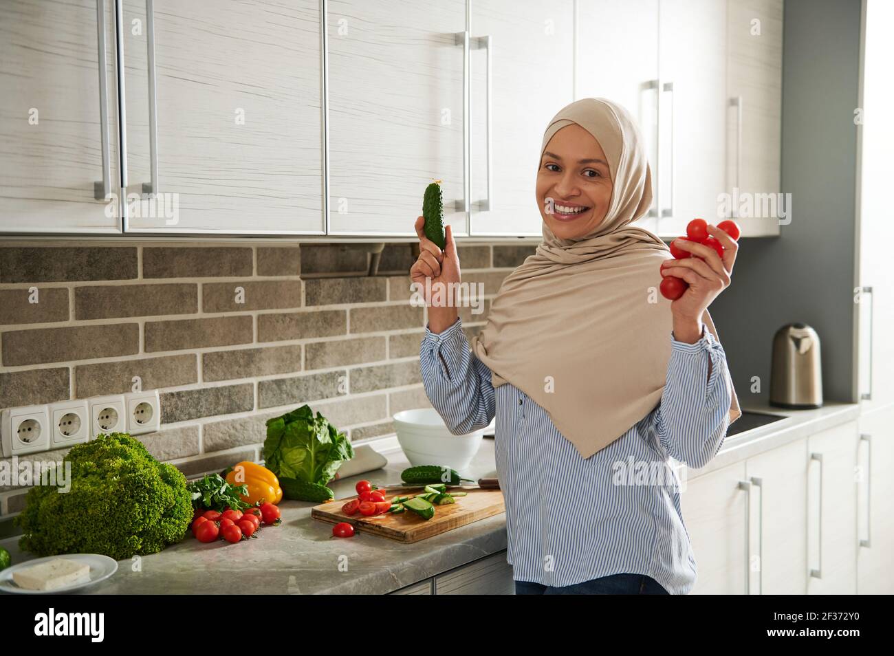 Smiling Muslim woman in hijab shows a cucumber and tomatoes to camera ...