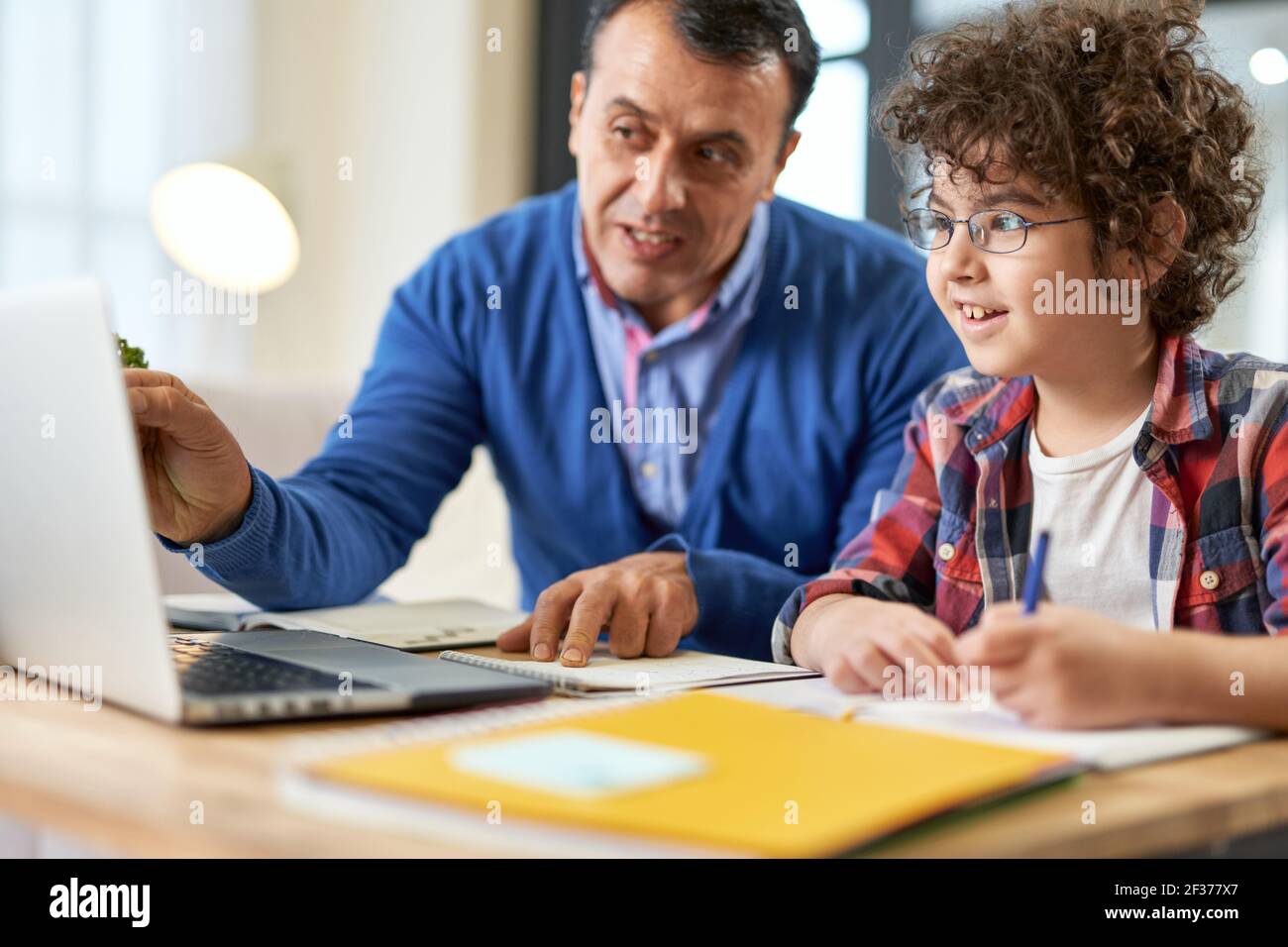 Smiling latin boy wearing glasses sitting at the desk together with his ...