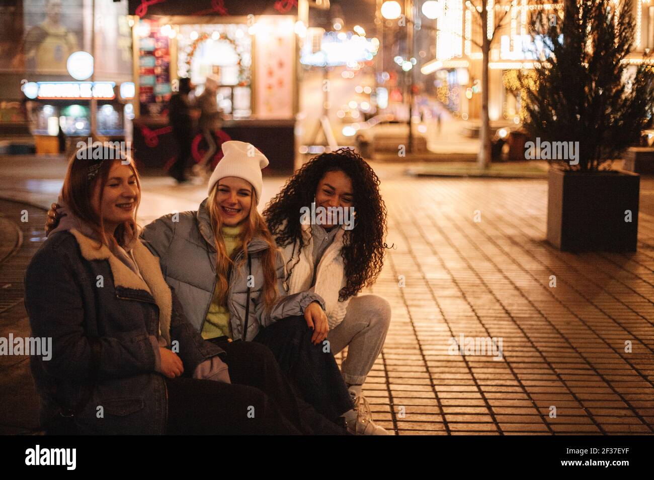 Happy female friends sitting on street in city at night Stock Photo - Alamy