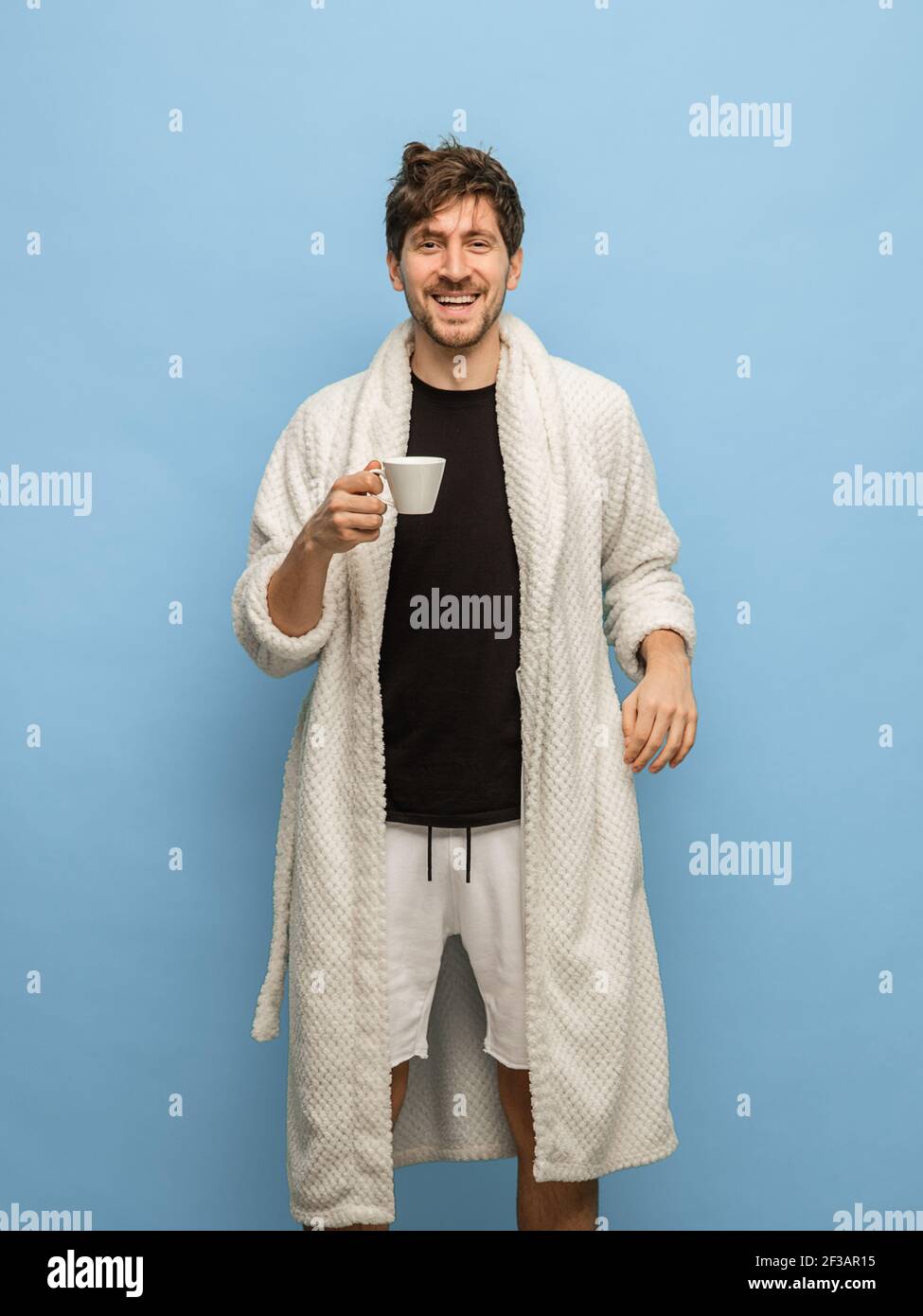 Happy young man in white bathrobe and cup of coffee isolated on blue ...