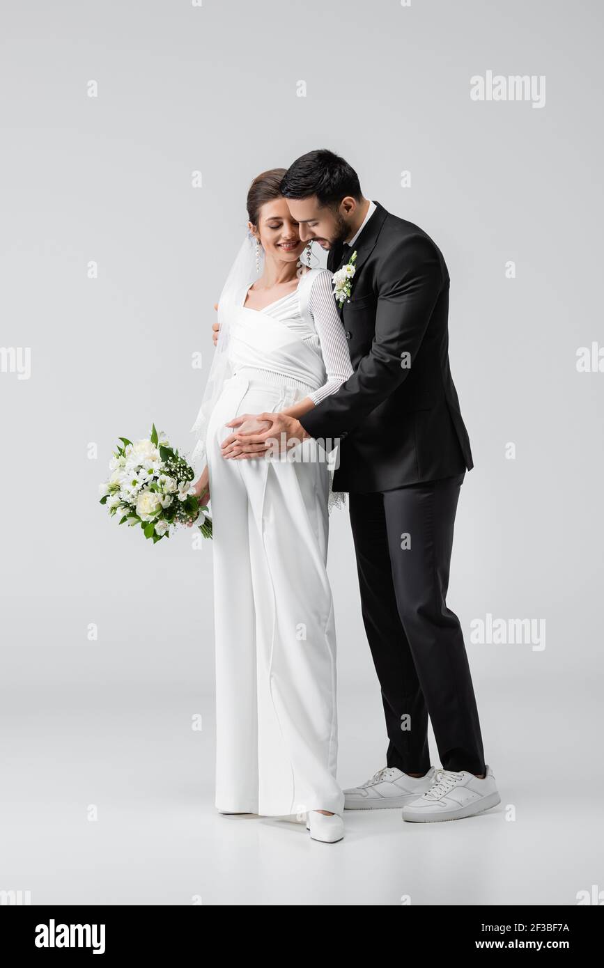Arabian groom hugging pregnant bride with bouquet on grey background ...