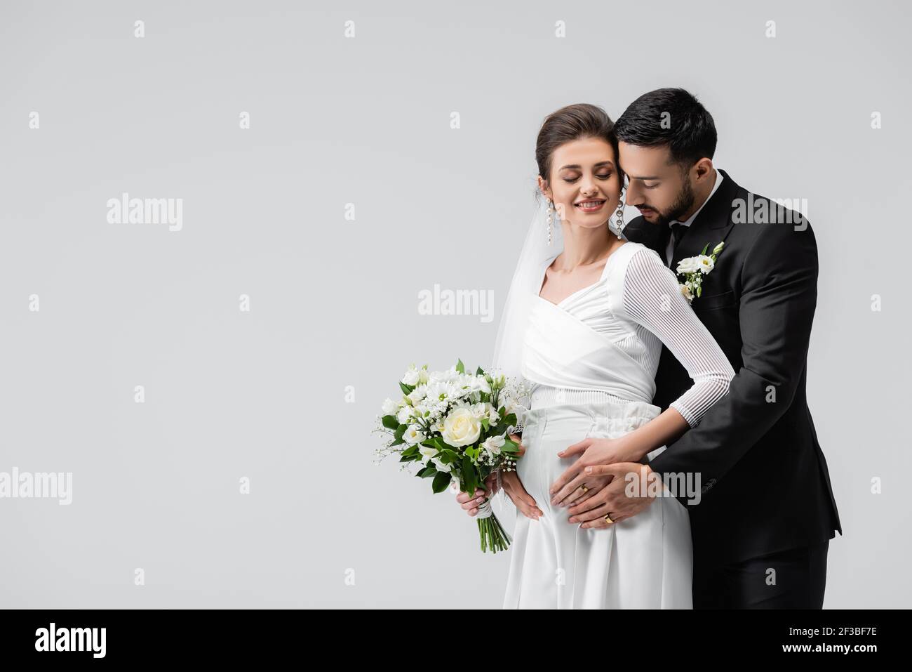 Muslim groom in suit hugging smiling pregnant bride isolated on grey ...