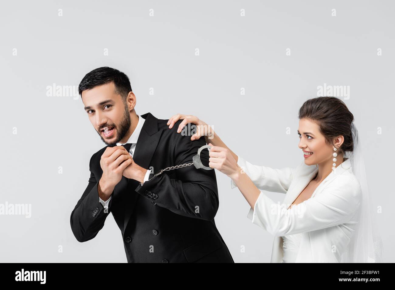 Smiling bride holding handcuffs of scared muslim groom isolated on grey ...