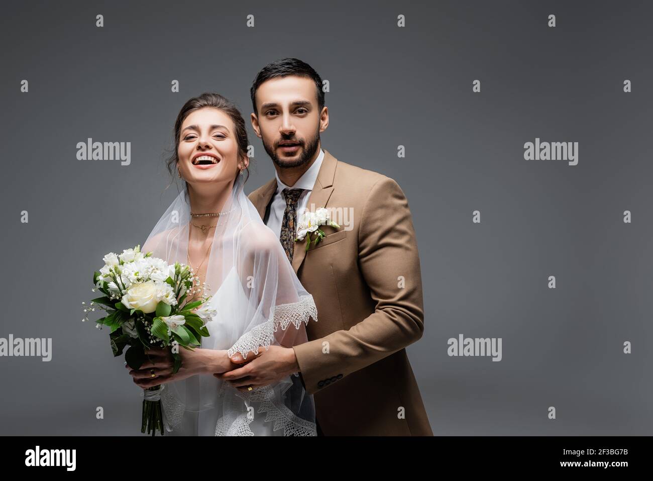 Arabian groom embracing cheerful bride with bouquet isolated on grey ...