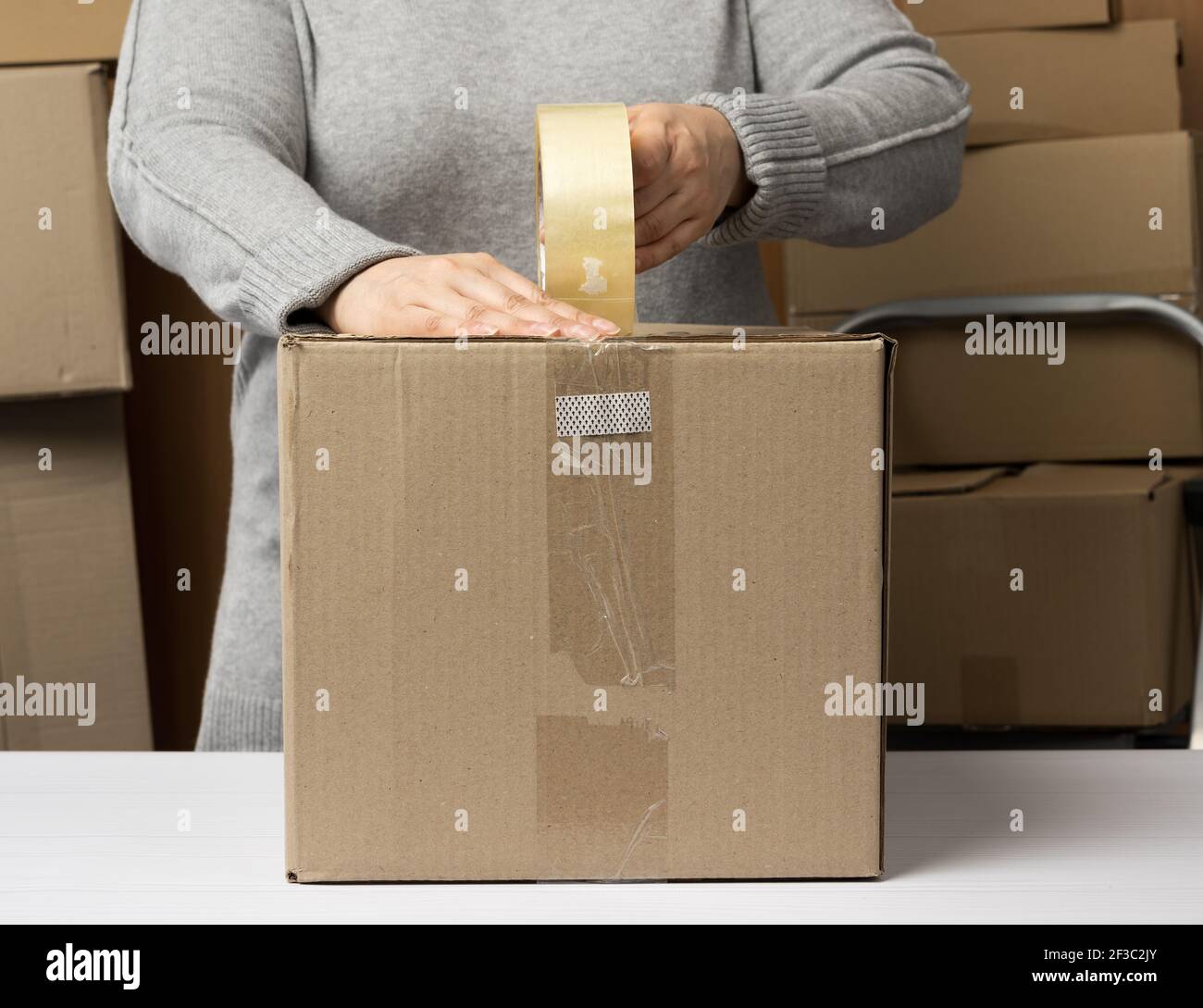 woman in a gray sweater holds a roll of duct tape and packs brown ...