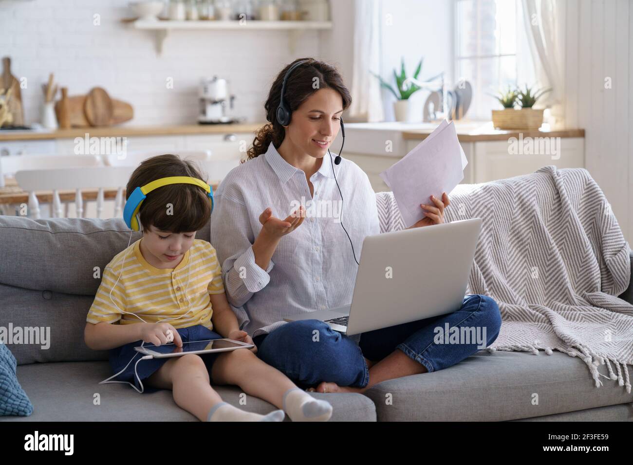 Freelancer mom sitting on sofa at home office during lockdown work on laptop, kid playing at ...
