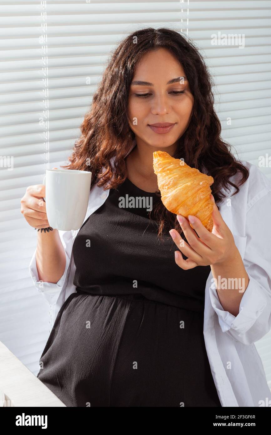 Happy pregnant young beautiful woman eating croissant during morning ...