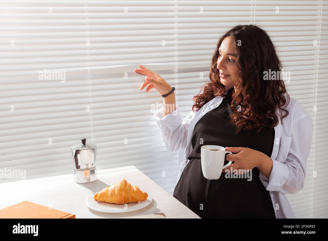 Joyful pregnant young beautiful woman looks through blinds during her ...
